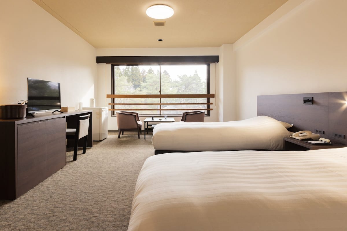 Quarto de hotel com duas camas, uma escrivaninha com televisão, uma mesinha e uma janela com vista para árvores.