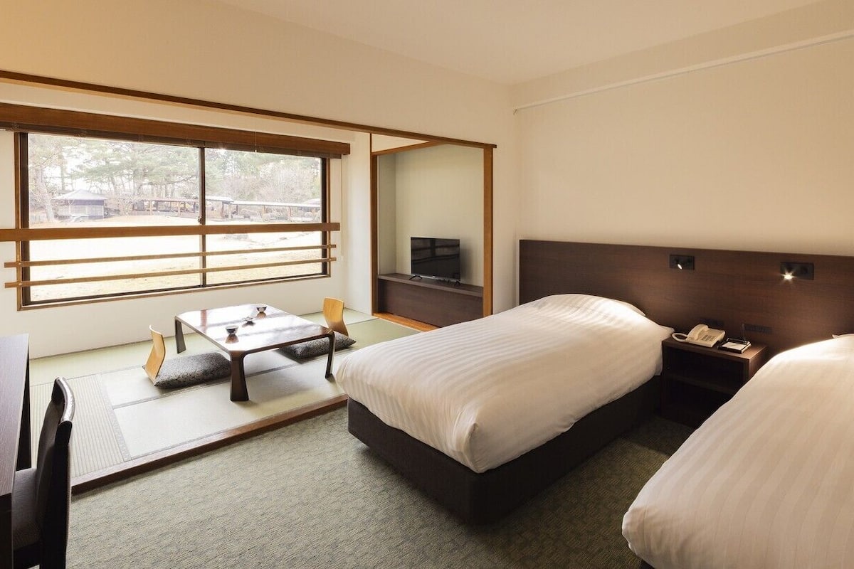 Quarto de hotel com duas camas, televisão, escrivaninha e janela com persianas.