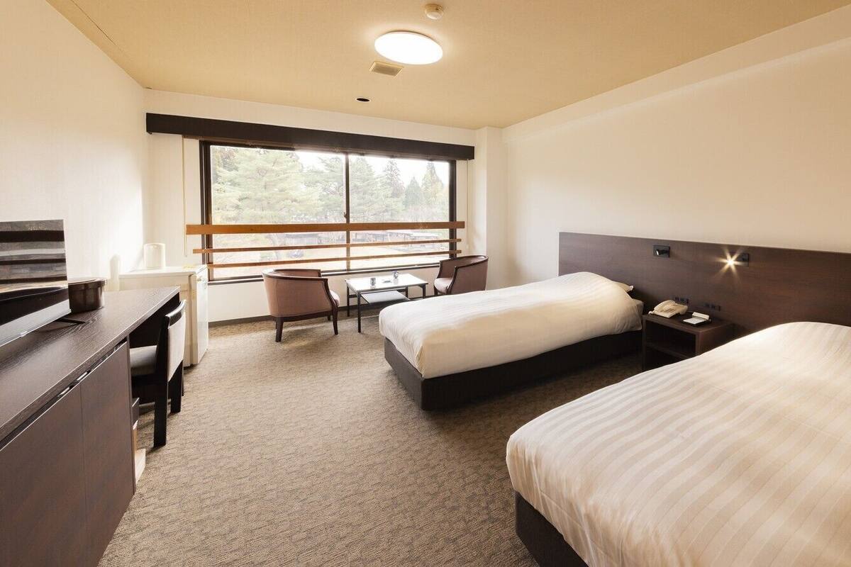 Quarto de hotel com duas camas, uma escrivaninha, uma cadeira, um armário e janela com vista para árvores.