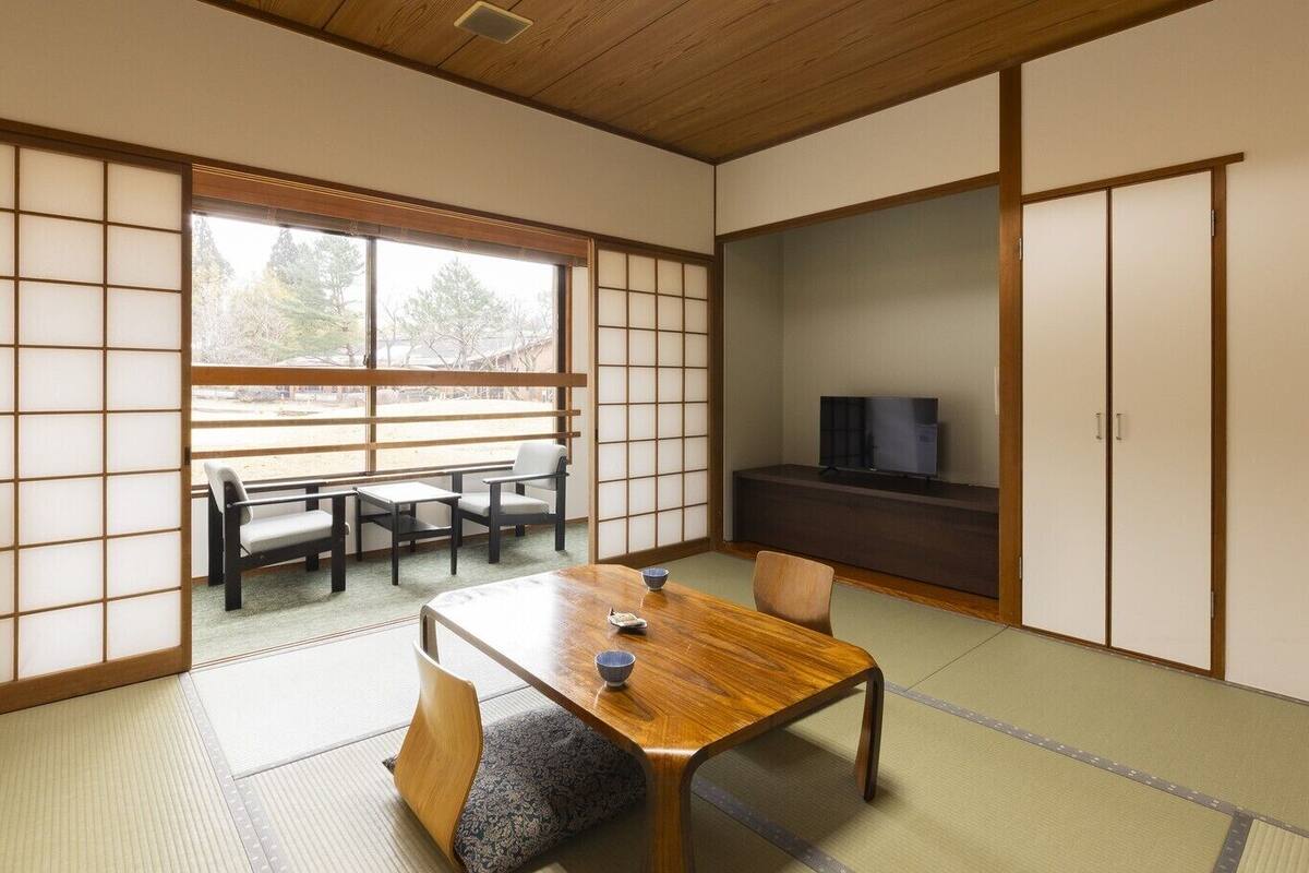 Um quarto japonês tradicional com piso de tatame, uma mesa baixa com almofadas, uma TV e portas deslizantes.