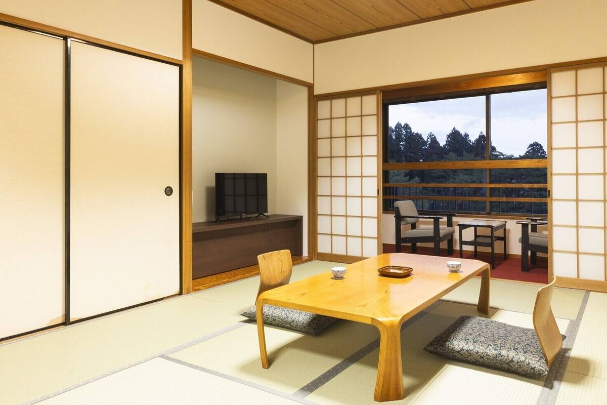 Um quarto japonês tradicional com tatames, uma mesa baixa com almofadas, uma televisão sobre um suporte e vista para árvores através de um biombos shoji.