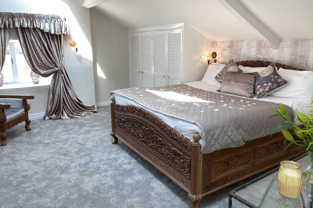 Quarto com cama grande, cadeira de madeira, janela com cortinas e luminária fixa na parede.