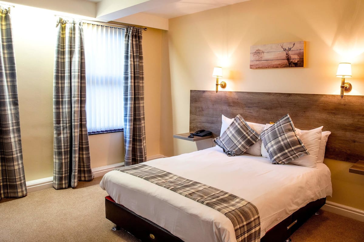 Quarto de hotel com uma cama grande, cortinas xadrez, cabeceira de madeira, duas luminárias de mesa e um quadro na parede.