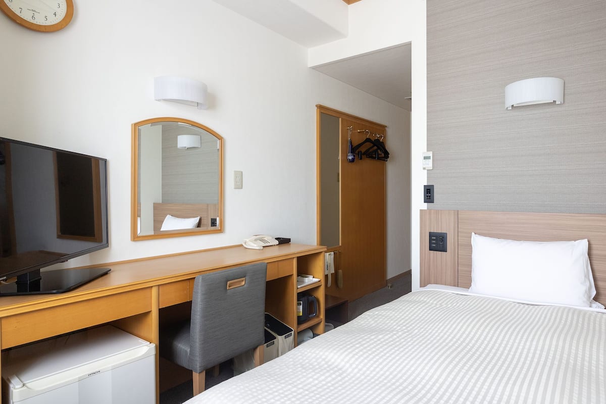 Quarto de hotel com cama, escrivaninha com computador, cadeira, televisão, espelho e armário.