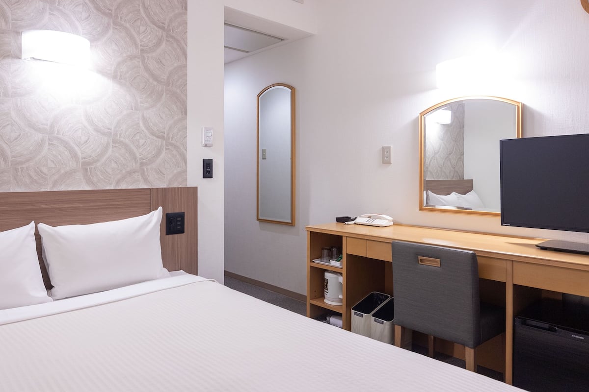 Quarto de hotel com cama, escrivaninha, cadeira, espelho e televisão.