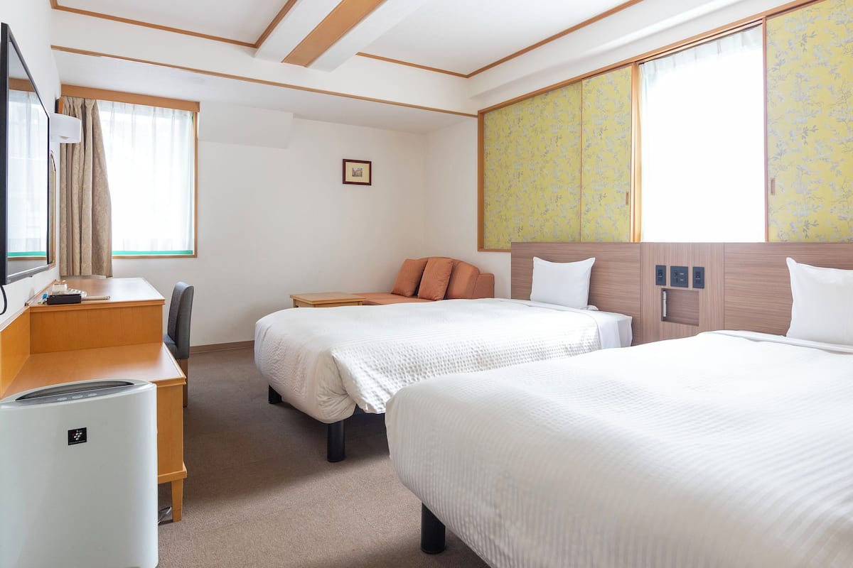 Quarto de hotel com duas camas, uma escrivaninha, uma cadeira, uma mesinha, um aquecedor e uma janela com cortinas.