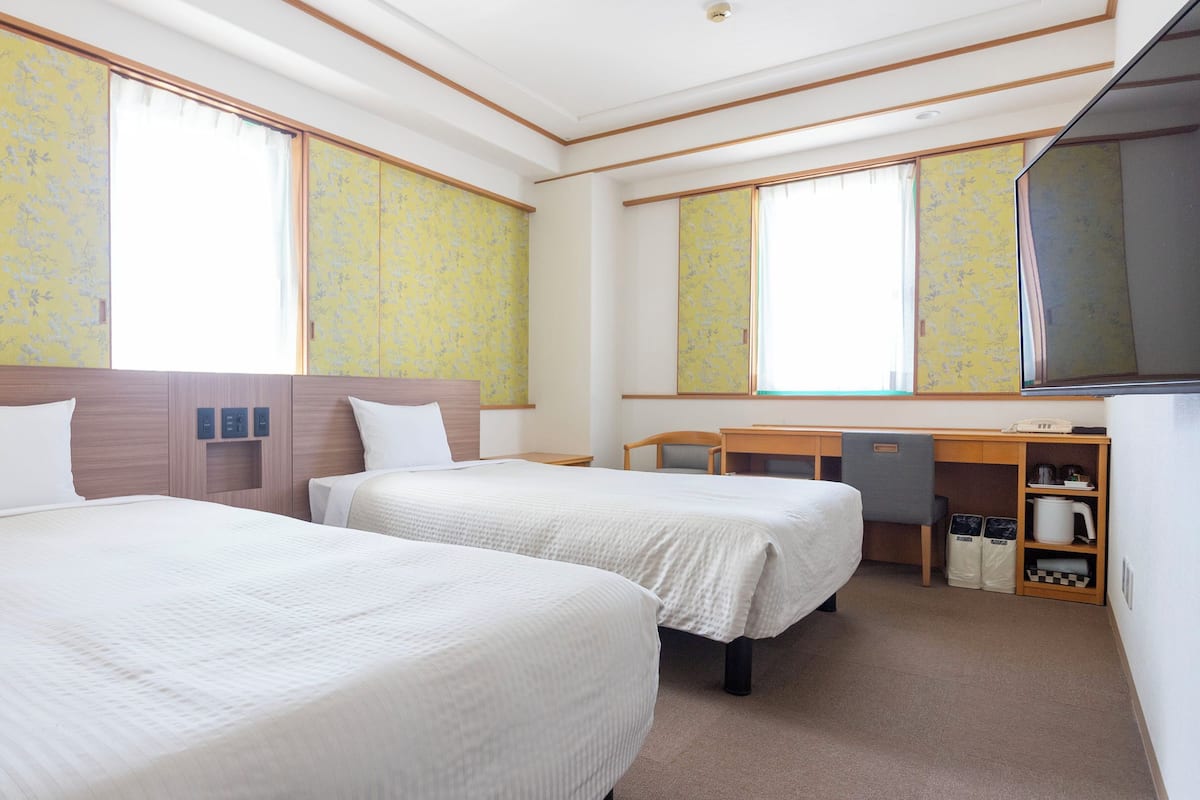 Quarto de hotel com duas camas, uma escrivaninha, uma cadeira, uma televisão e um guarda-roupa.
