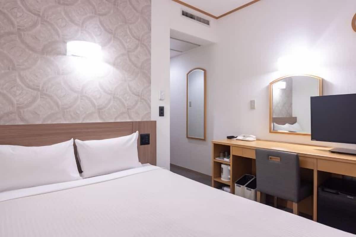 Quarto de hotel com uma cama, uma mesa com computador, um espelho e uma luminária na parede.
