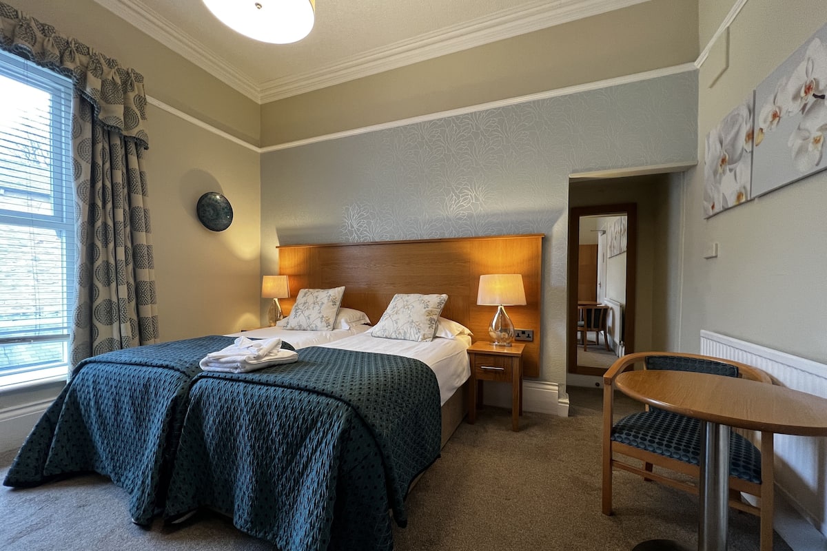 Quarto de hotel com uma cama grande, duas luminárias de cabeceira, uma mesinha e uma cadeira.