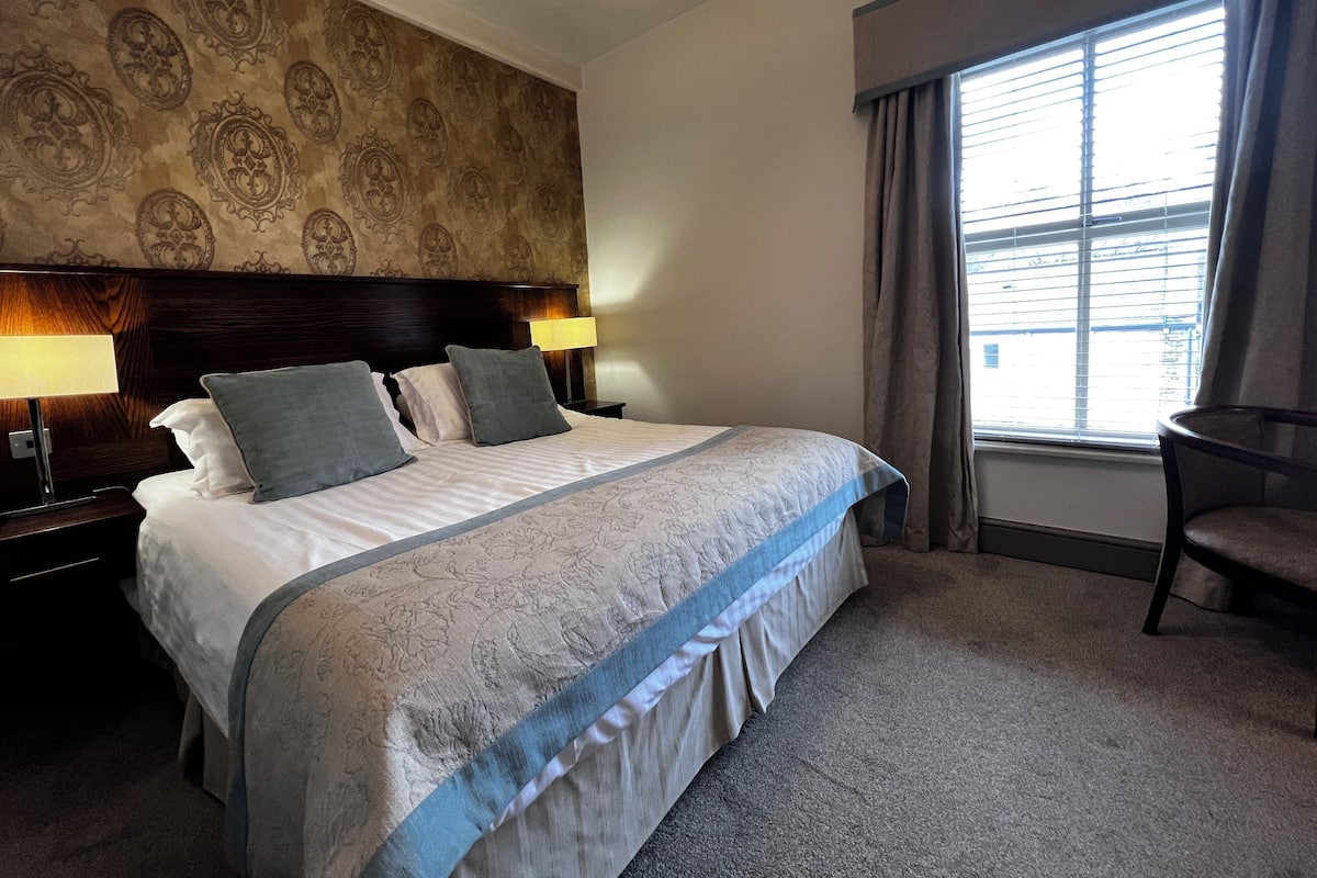 Quarto de hotel com uma cama grande, duas luminárias de cabeceira, cabeceira com papel de parede estampado, janela com cortinas e uma cadeira.