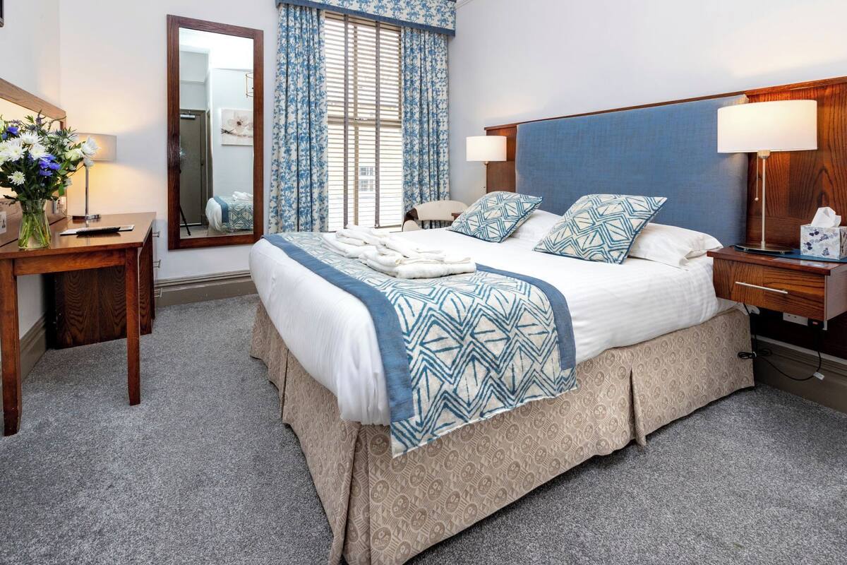 Quarto de hotel com uma cama grande, uma mesa de cabeceira com abajur, um espelho e uma janela com cortinas azuis.