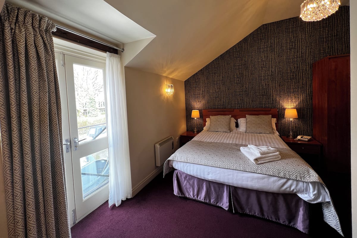 Quarto de hotel com uma cama, duas mesas de cabeceira, um candelabro, uma janela com cortinas e parede estampada.