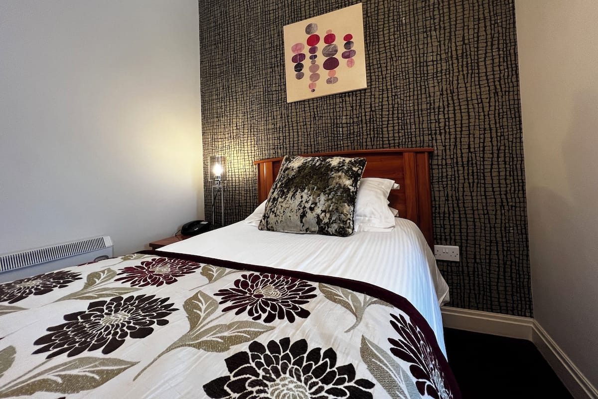 Quarto de hotel com uma cama grande, cabeceira, obra de arte na parede, luminária de cabeceira e edredom com padrão floral.