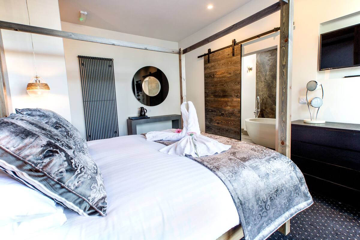 Quarto de hotel com cama, mesa de cabeceira, espelho, porta de madeira e televisão presa à parede.
