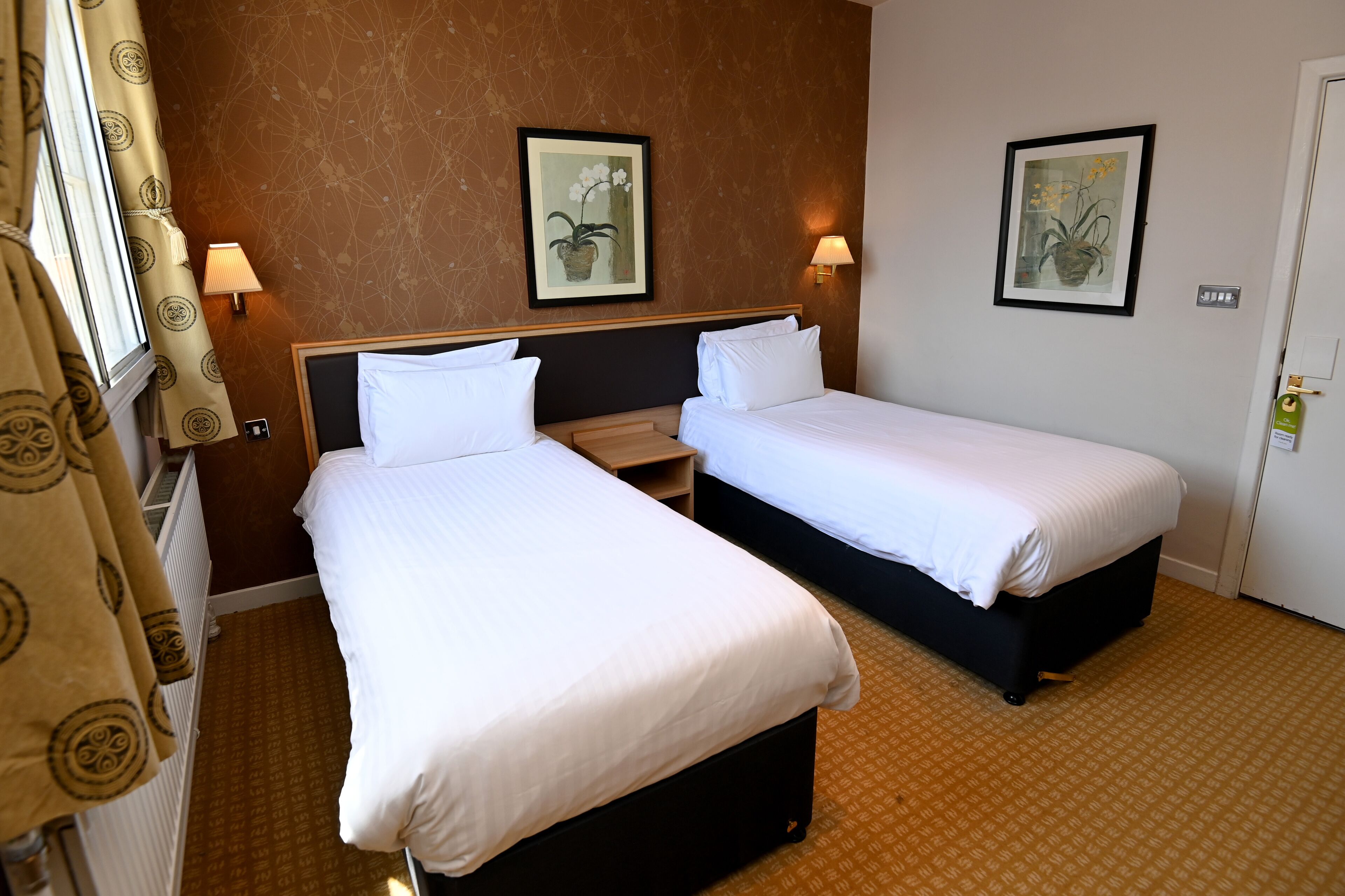 Quarto de hotel com duas camas de solteiro, cabeceira de madeira, mesinha de cabeceira pequena, luminária fixada na parede e dois quadros florais emoldurados.