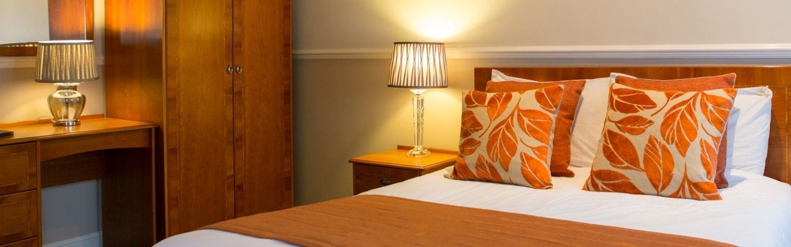 Quarto de hotel com cama de madeira, duas almofadas estampadas com folhagens, abajur e guarda-roupa de madeira.
