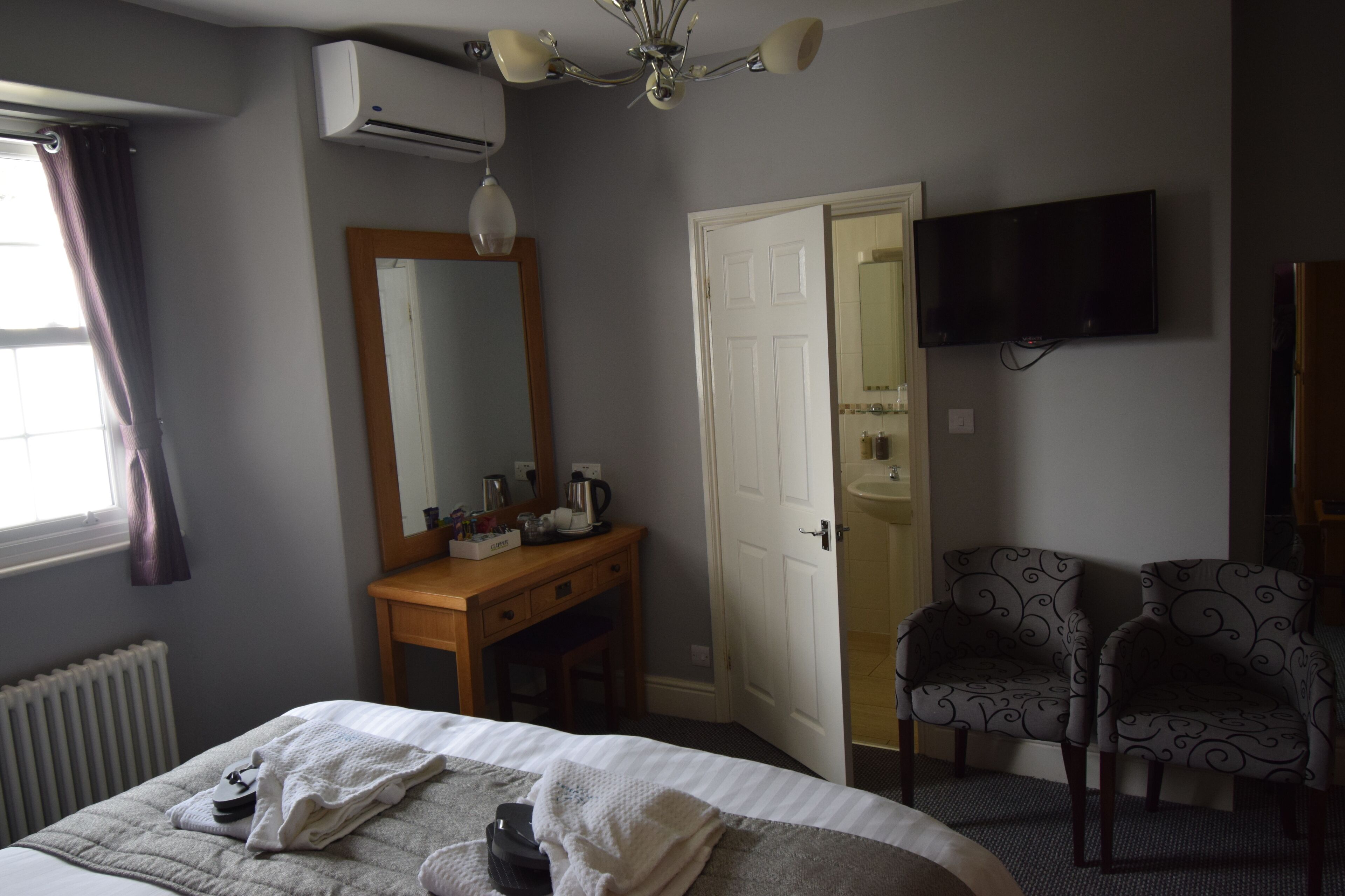 Quarto de hotel com cama, escrivaninha com espelho, cadeira, televisão e ar condicionado.