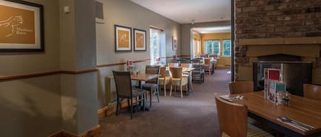 Uma sala de jantar de hotel com várias mesas e cadeiras, obras de arte nas paredes e uma lareira de tijolos.