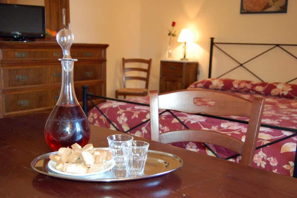 Quarto com cômoda de madeira, cama com colcha florida, mesa de cabeceira com abajur e bandeja com comida e bebidas sobre uma mesa.