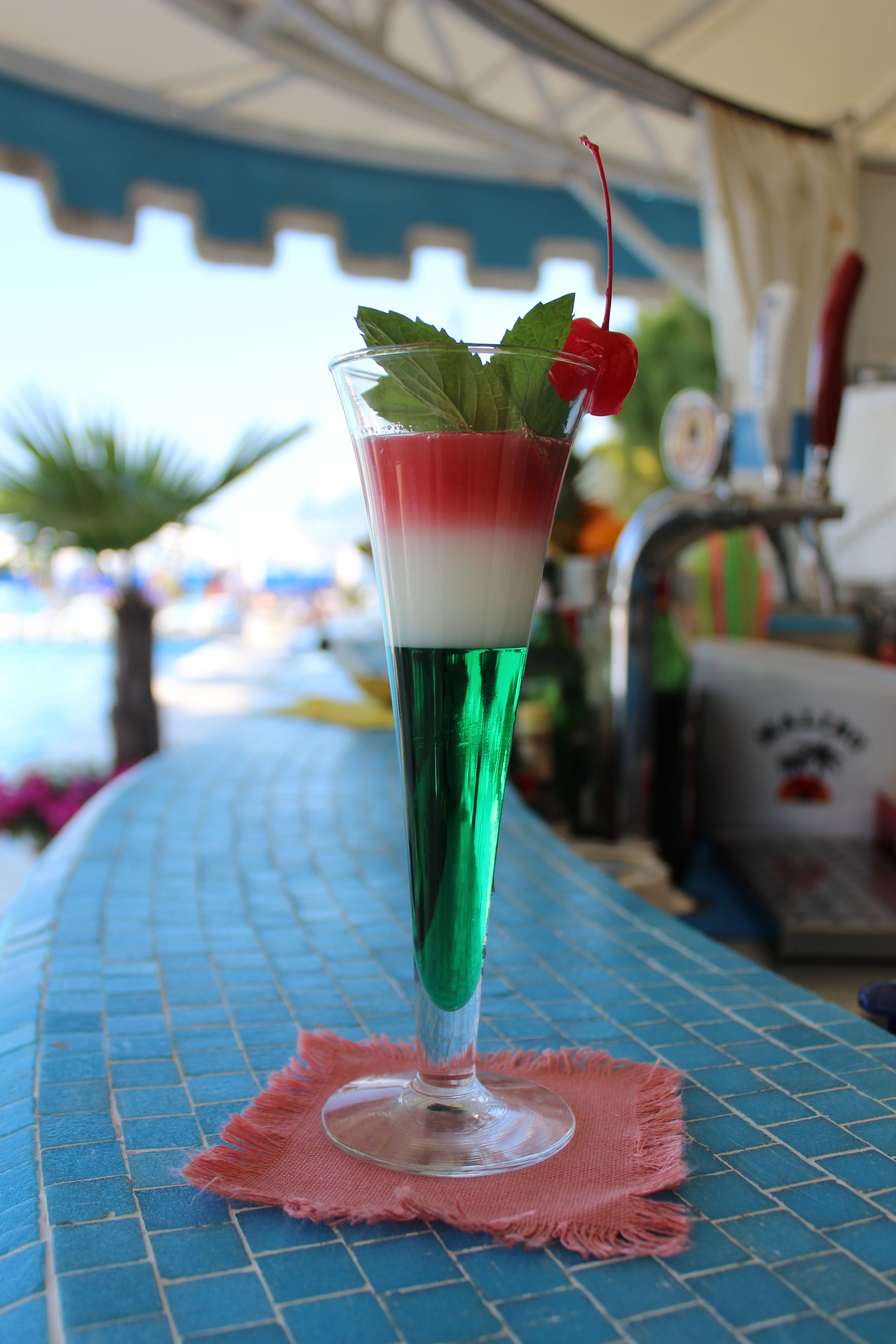Um copo de cocktail com bebida em camadas, decorado com uma cereja e folhas de hortelã, sobre uma toalha de mesa azul com um apoio de mesa cor-de-rosa.