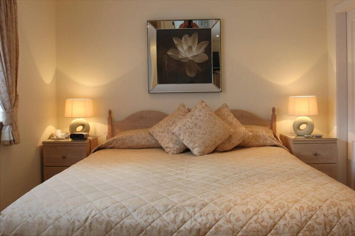 Uma cama bem arrumada com colcha estampada de flores, duas mesas de cabeceira com abajures, e uma obra de arte emoldurada sobre a cama.