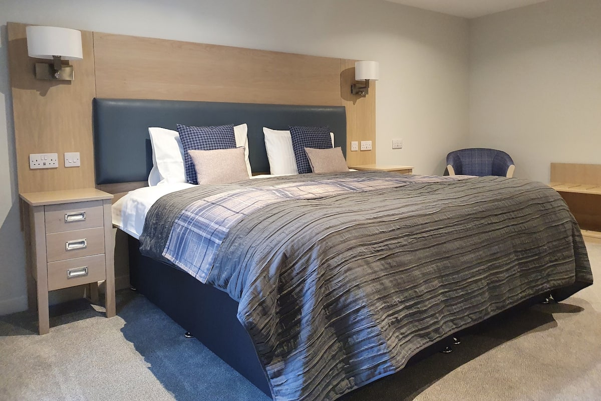 Quarto de hotel moderno com uma cama grande, mesas de cabeceira, escrivaninha e uma cadeira.