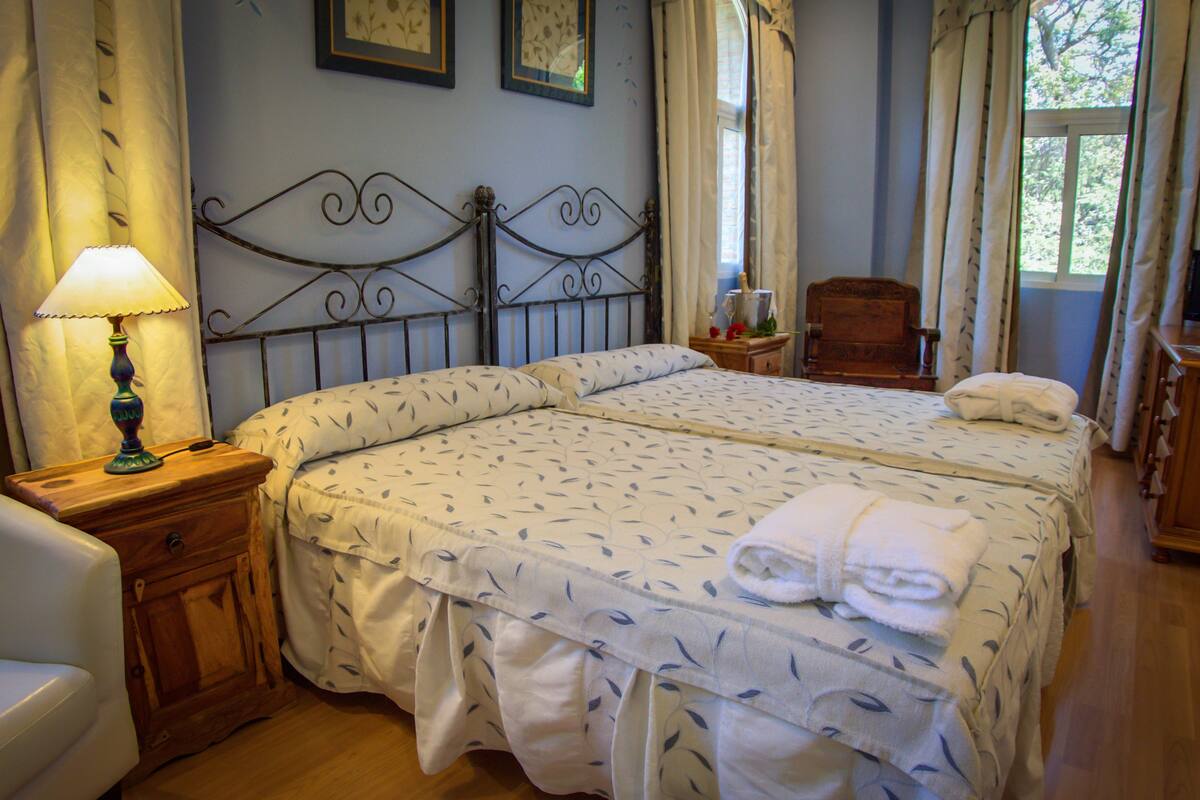Uma cama de casal com colcha estampada em folhas, um criado-mudo de madeira com abajur e uma cadeira de madeira.