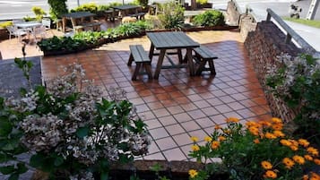 Uma área de pátio ao ar livre com piso de azulejos, mesas de piquenique de madeira e diversas plantas e flores.