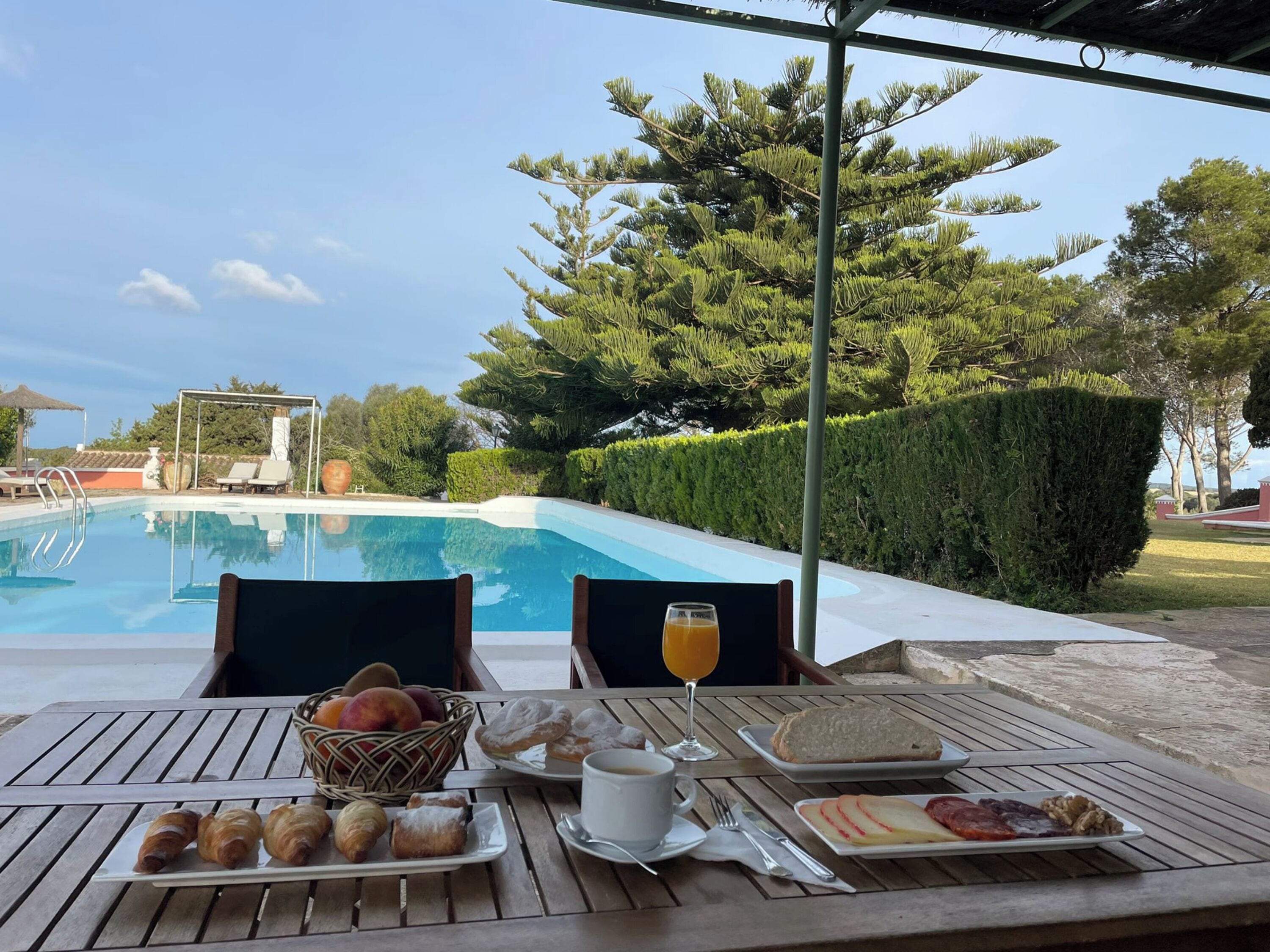 Mesa à beira da piscina com itens de café da manhã, um copo de suco de laranja e vista para a piscina e vegetação ao redor.