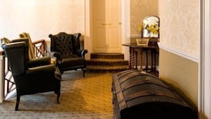 Um quarto com carpete estampado, um banco de madeira, uma poltrona escura revestida e uma mesa pequena com um vaso de flores.