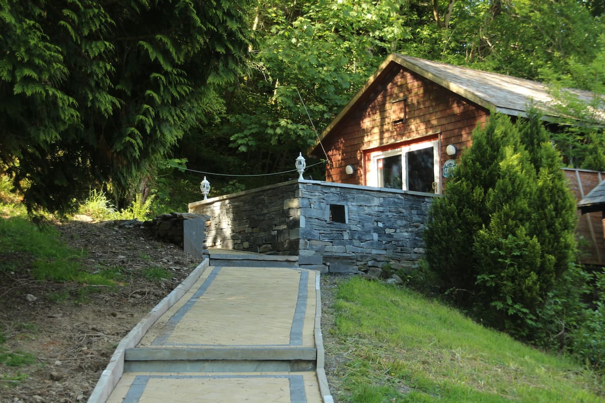 Uma cabana rústica com base de pedra e telhado de madeira, rodeada por uma floresta densa.