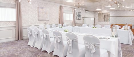 Uma sala de banquetes com mesas e cadeiras brancas, lustres e piso coberto de carpete.