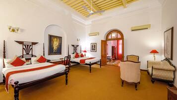 Quarto de hotel com duas camas, área de estar com cadeiras, ventilador de teto e uma porta arqueada que dá para outro cômodo.