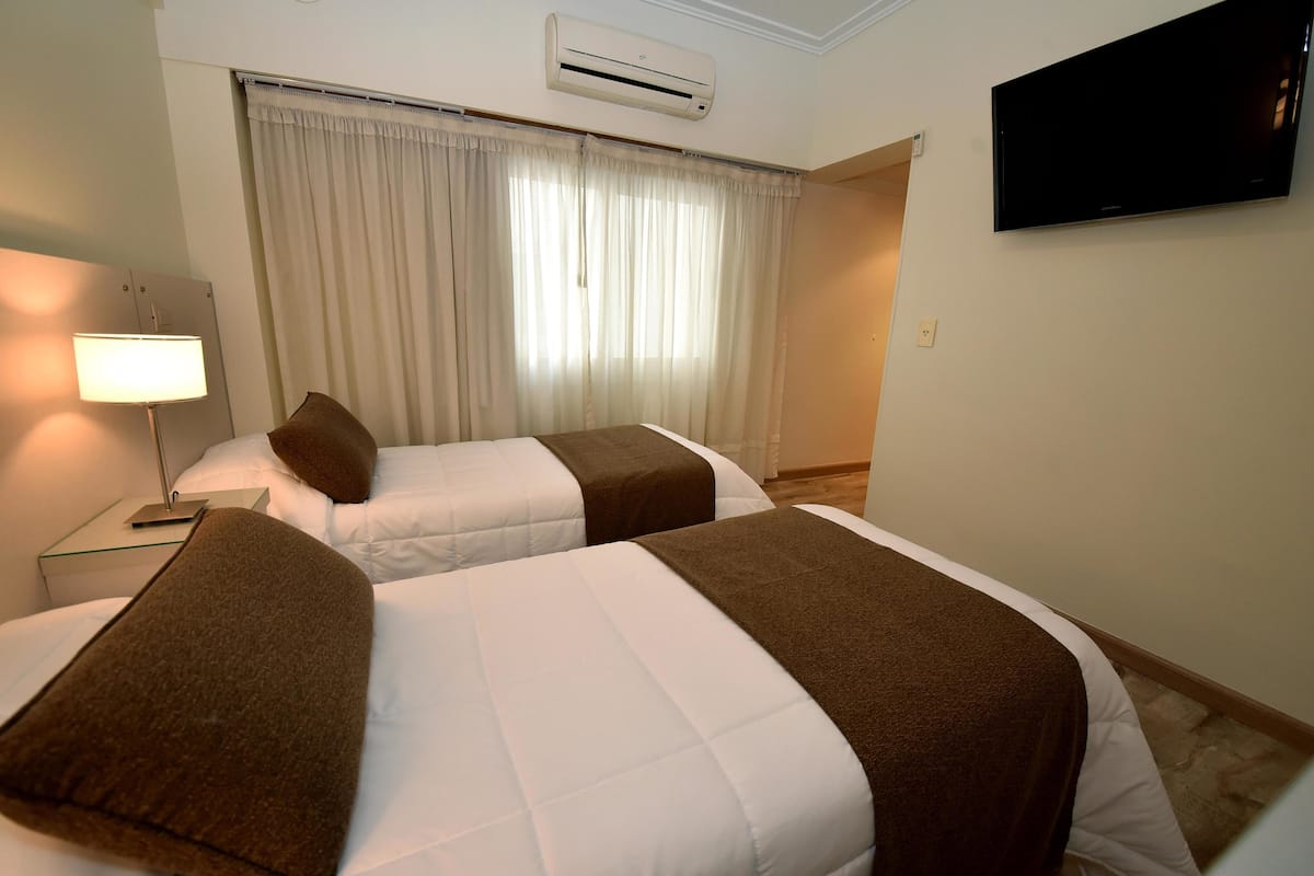 Quarto de hotel com duas camas, televisão de tela plana, aparelho de ar condicionado, luminária e janela com cortinas.