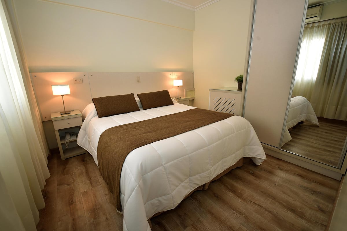 Quarto de hotel com cama grande, luminárias laterais, mesa de cabeceira, espelho e piso de madeira.