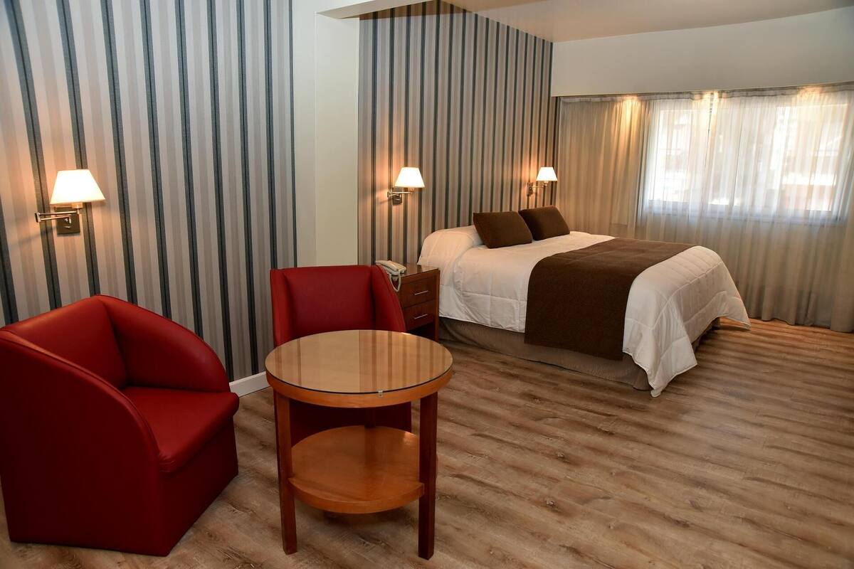 Quarto de hotel com cama, poltrona vermelha, mesa de madeira pequena e papel de parede listrado.