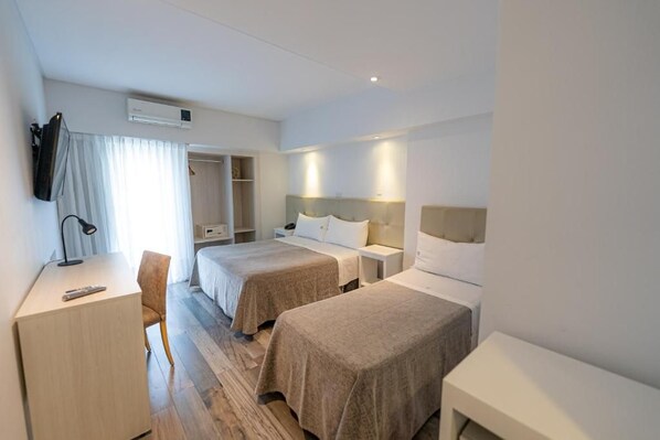 Quarto de hotel com duas camas, uma escrivaninha, uma cadeira, guarda-roupa e ar condicionado.