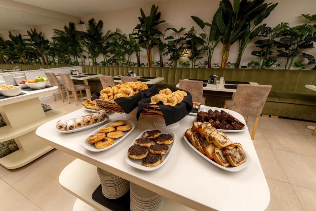 Mesa de buffet com diversos pães e doces, rodeada por cadeiras e mesas em um ambiente com plantas tropicais ao fundo.