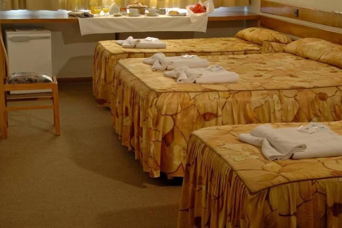 Quarto de hotel com três camas, cada uma com colcha estampada e toalhas dobradas com capricho. Uma pequena mesa com itens de café da manhã é visível ao fundo.