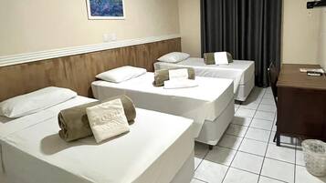 Quarto de hotel com três camas de solteiro, cabeceira de madeira, televisão de tela plana, escrivaninha e uma mesinha.
