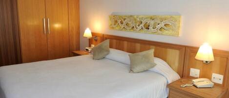 Quarto de hotel com uma cama grande, duas mesas de cabeceira, um guarda-roupa de madeira e um objeto decorativo na parede.