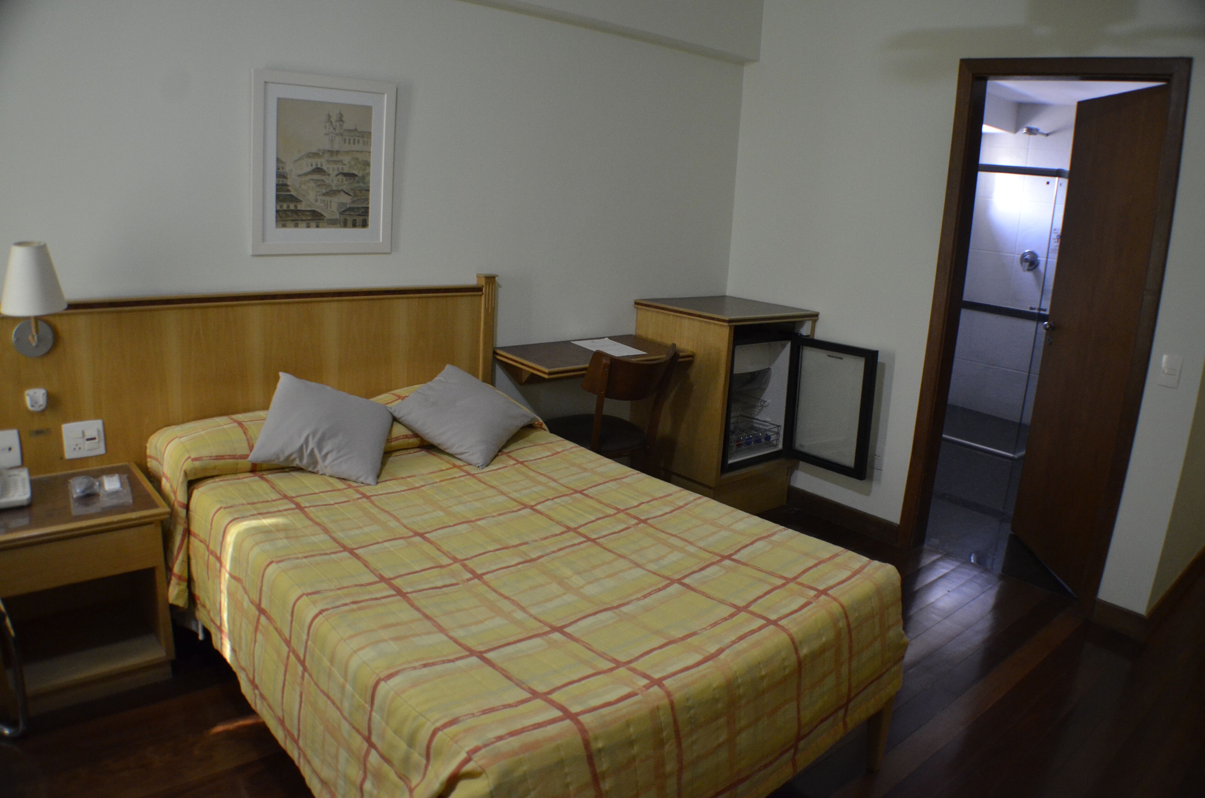 Quarto de hotel com cama, escrivaninha, cadeira, mesa de cabeceira, luminária e um quadro na parede.