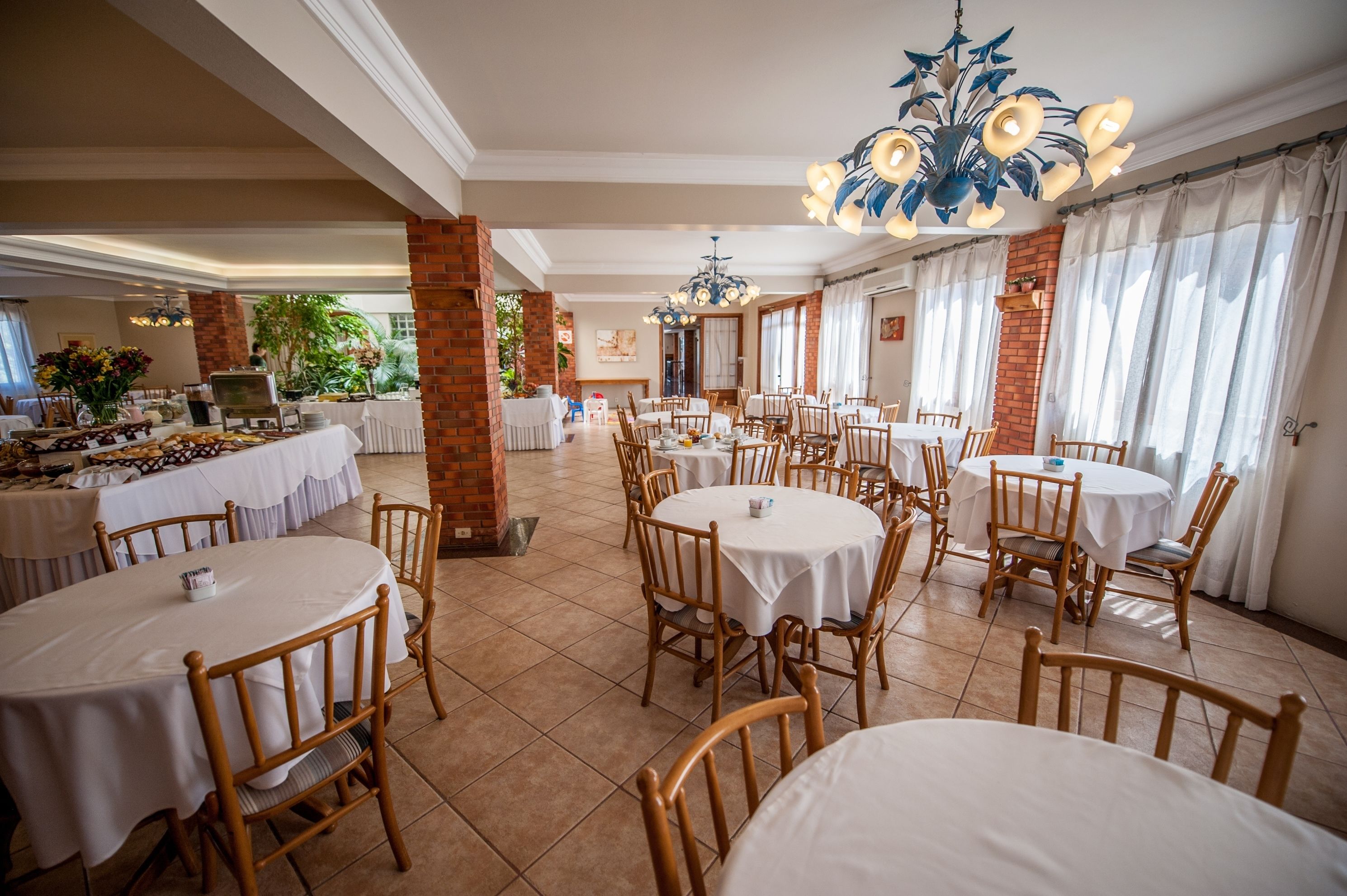 Uma sala de jantar ampla com mesas redondas e cadeiras de madeira, com um lustre e colunas de tijolo.