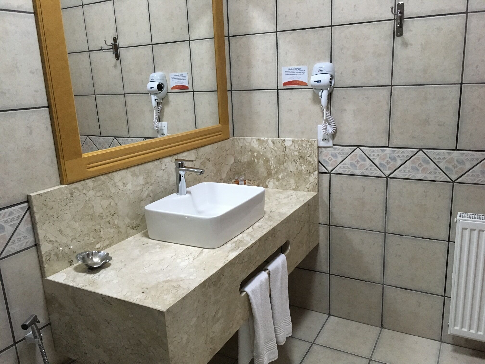 Uma casa de banho com uma pia retangular, um espelho e dois secadores de cabelo fixados na parede.
