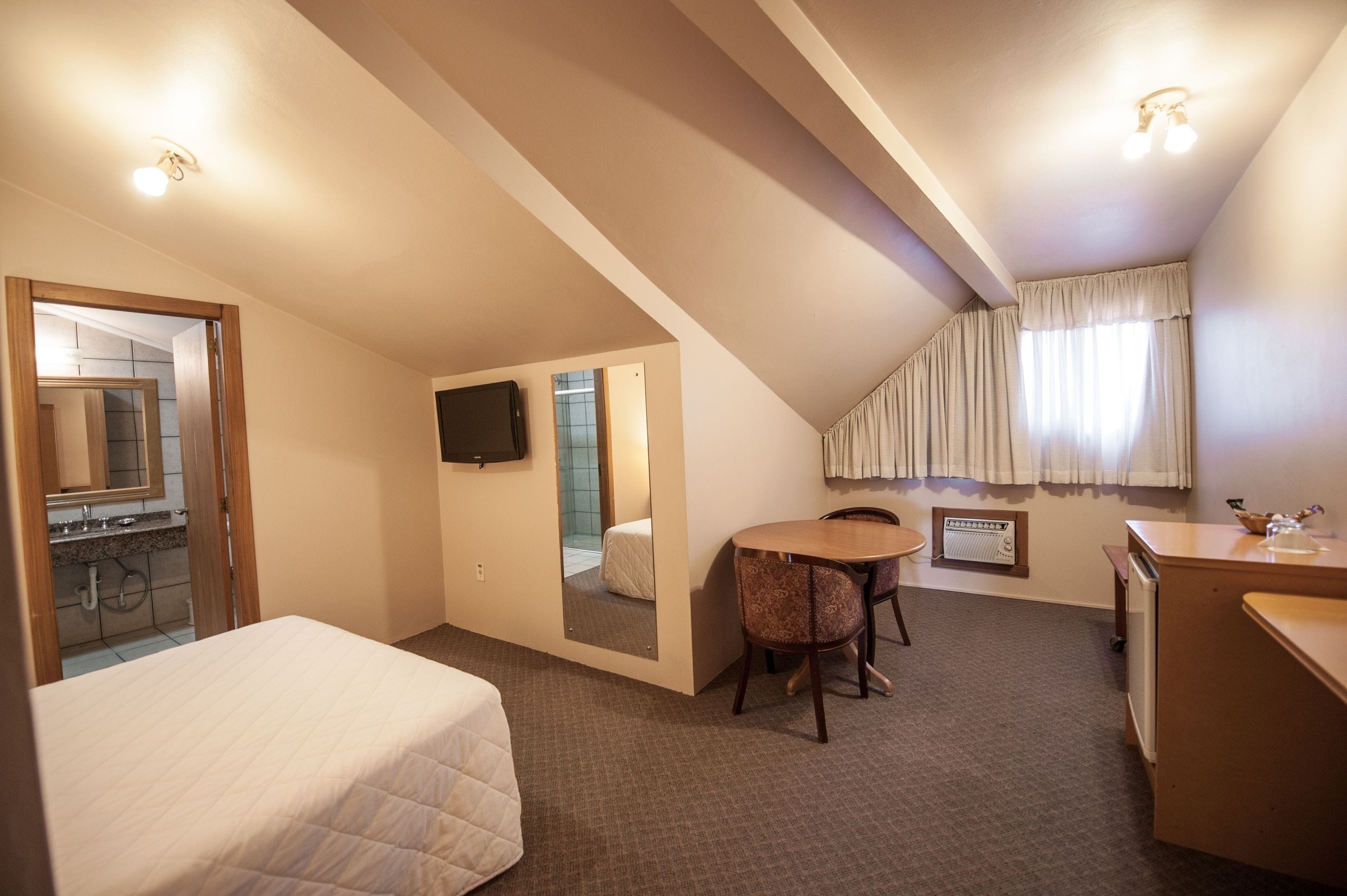 Quarto de hotel com cama, escrivaninha, cadeira, mesa pequena, espelho, janela com cortinas e televisão.