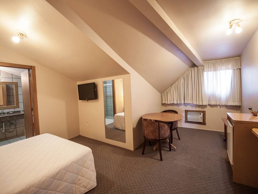 Quarto de hotel com cama, escrivaninha, cadeira, mesa pequena, espelho, janela com cortinas e televisão.