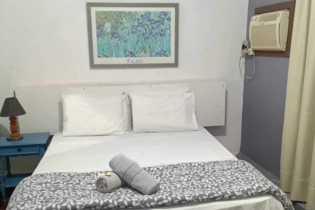 Cama bem arrumada com um edredom branco, manta cinza e branca estampada e toalha cinza enrolada. Uma obra de arte floral emoldurada está pendurada acima da cama.