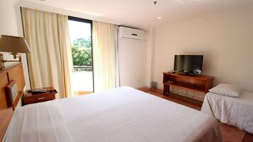 Quarto de hotel com uma cama grande, uma televisão sobre um suporte de madeira, uma escrivaninha com uma luminária e uma janela com cortinas.