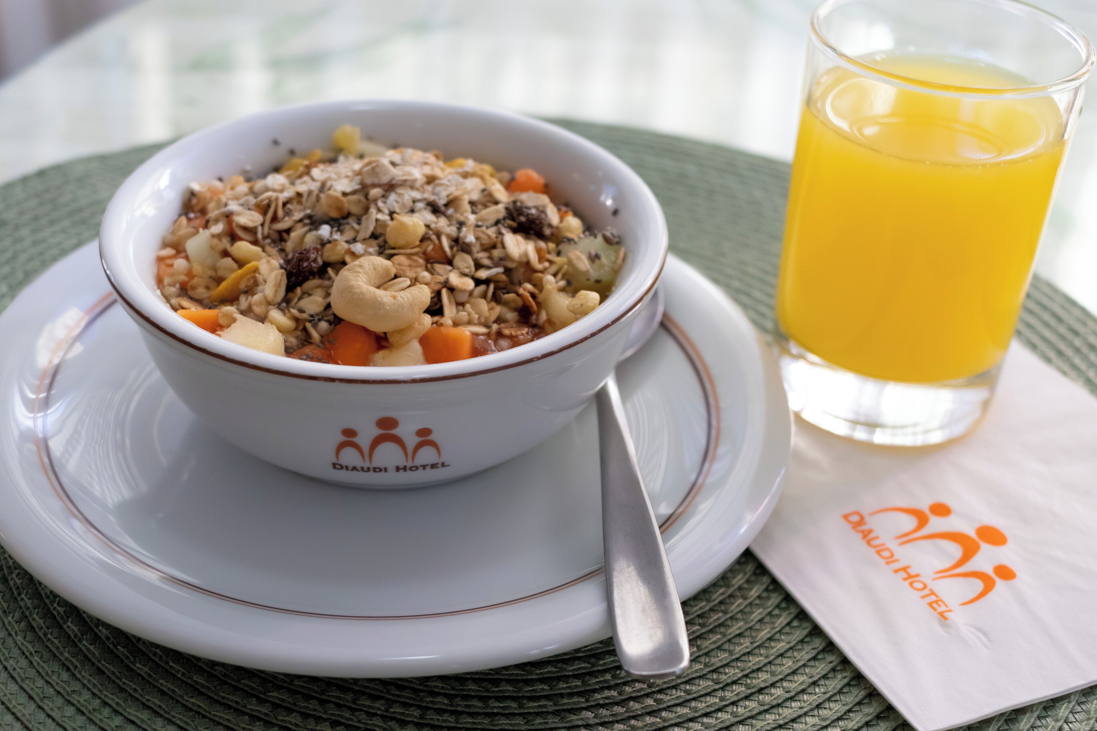 Uma tigela de cereais com um copo de suco de laranja sobre uma mesa, com um descanso de sousa e um guardanapo com o logotipo do hotel.