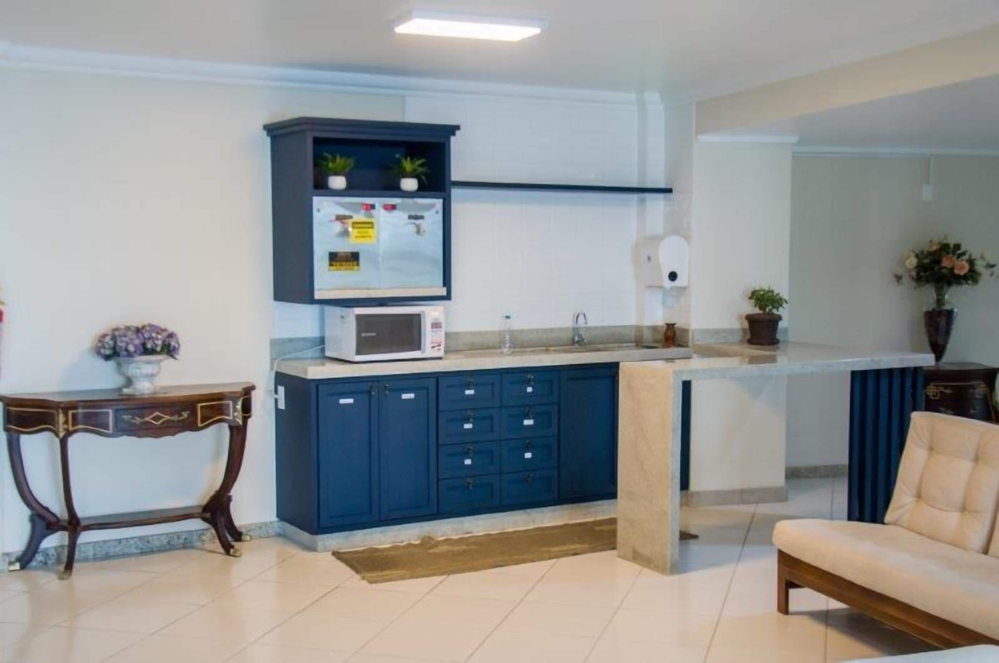 Uma cozinha compacta com micro-ondas, pia e um pequeno armário. Há uma mesa de madeira com um vaso de flores e um sofá na sala.